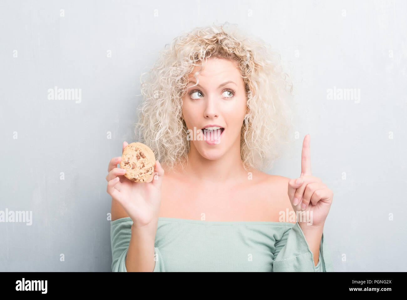 Young blonde woman over grunge grey wall eating chocolate chip cooky ...