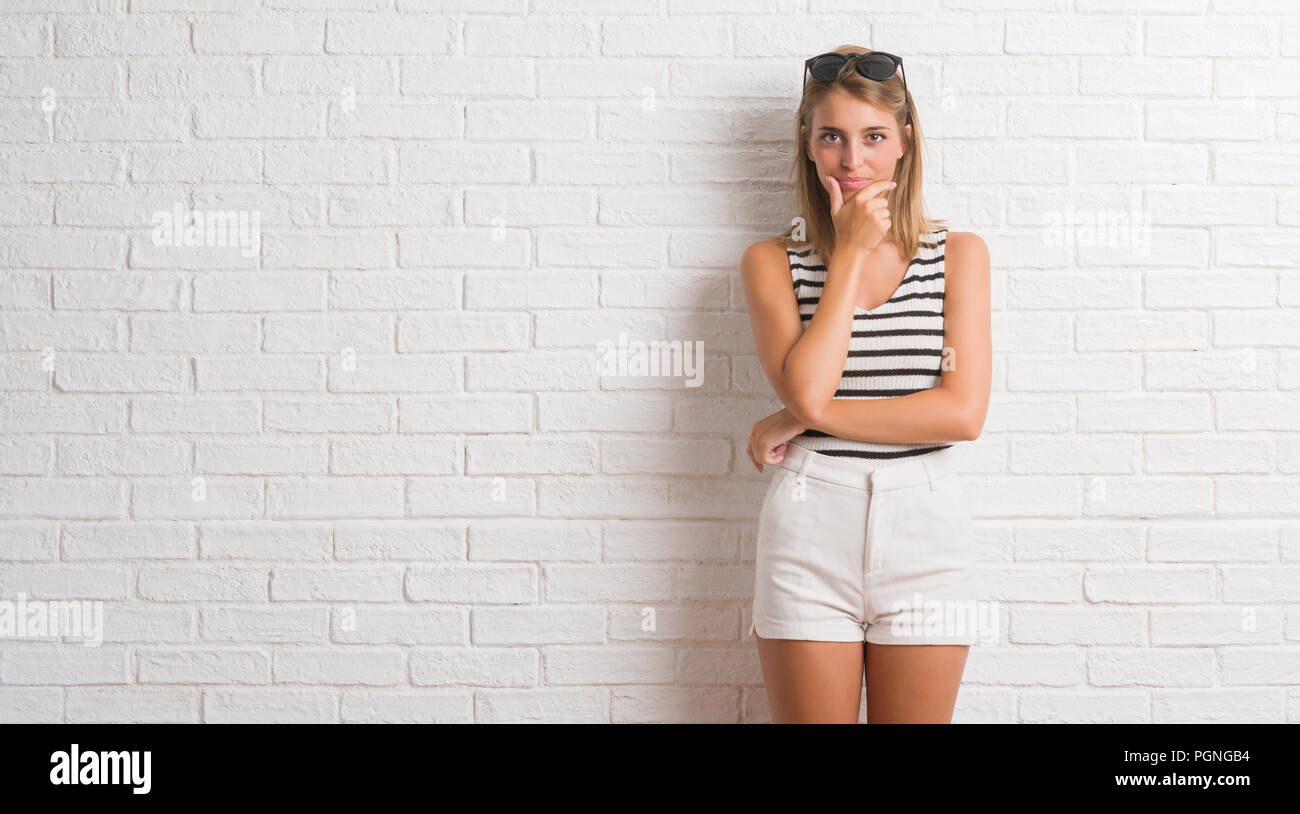 Young beautiful blonde woman over white brick wall looking confident at ...