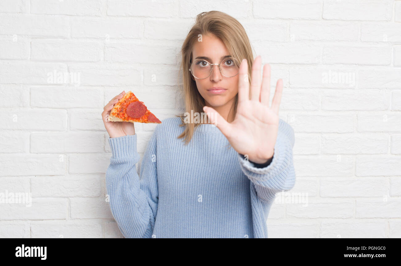 Beautiful young woman over white brick wall eating pizza slice with ...