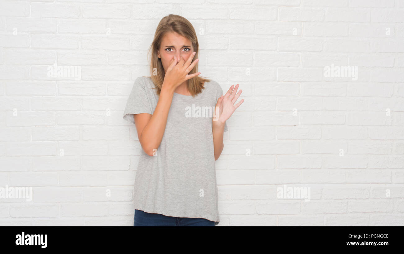 Beautiful young woman over white brick wall smelling something stinky ...