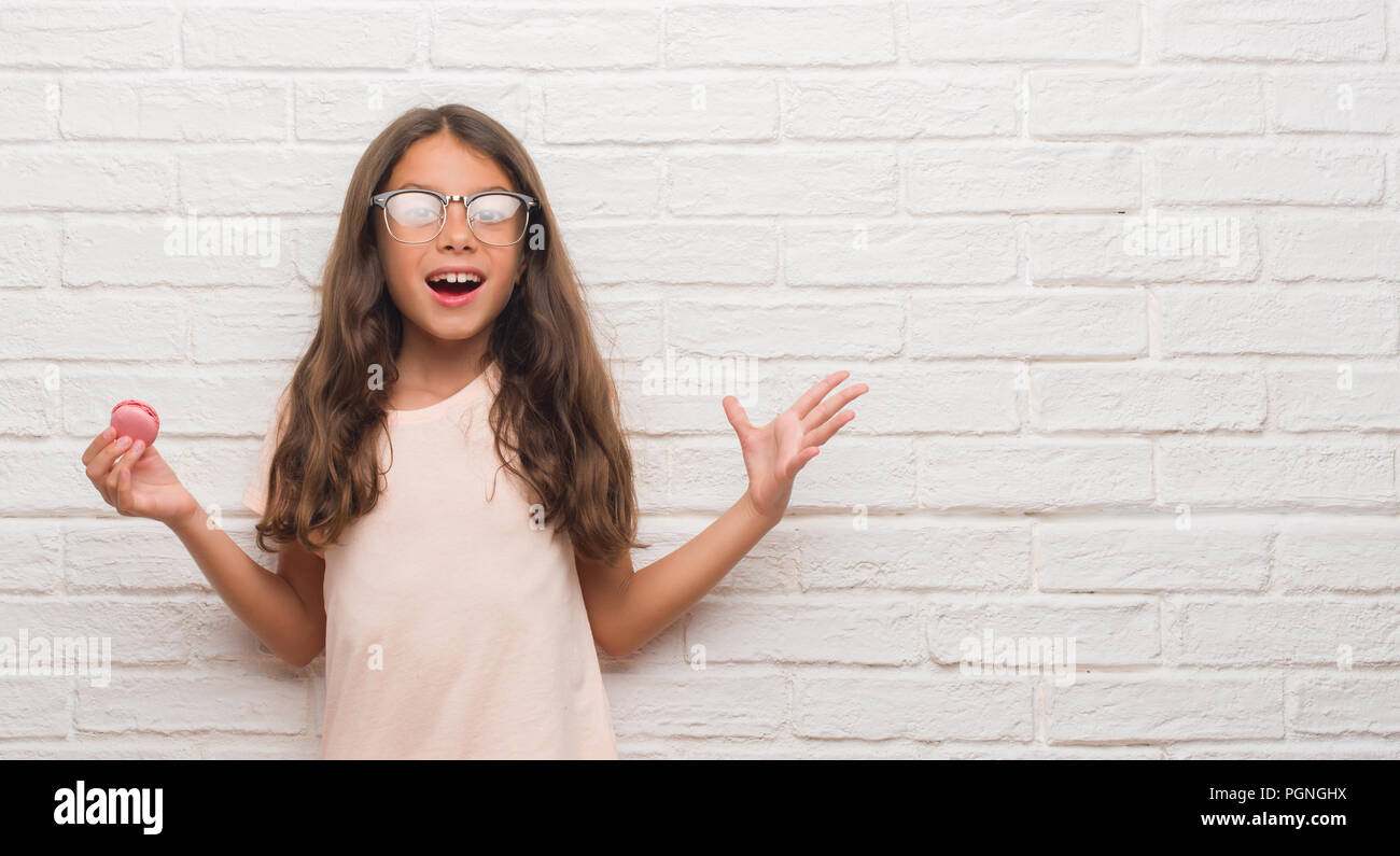 Young hispanic kid over white brick wall eating pink macaron very happy ...