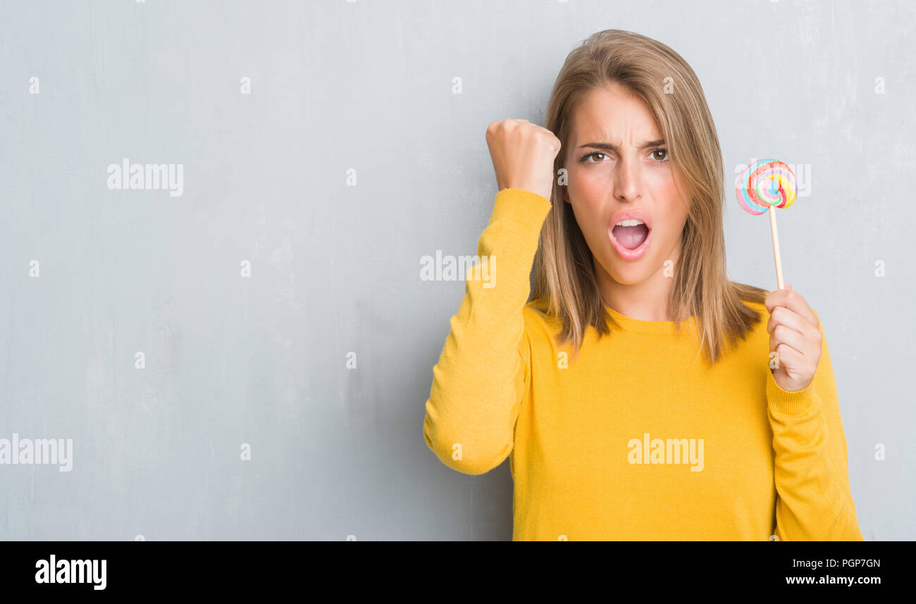 Beautiful young woman over grunge grey wall eating lollipop candy ...