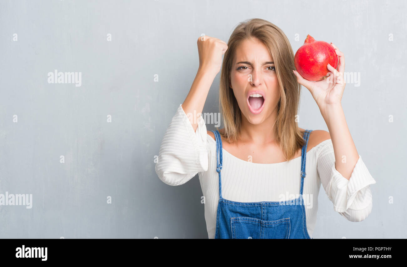 Beautiful young woman over grunge grey wall holding pomegranate annoyed ...