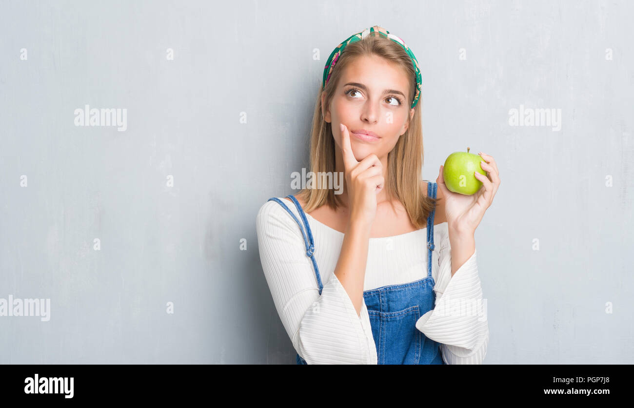 Beautiful young woman over grunge grey wall eating green apple serious ...