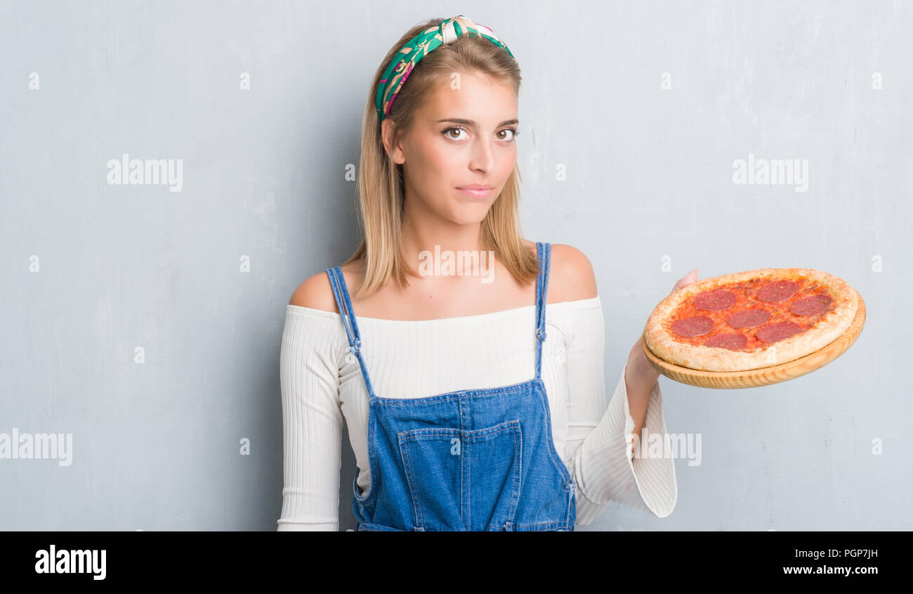 Beautiful young woman over grunge grey wall eating pepperoni pizza with ...