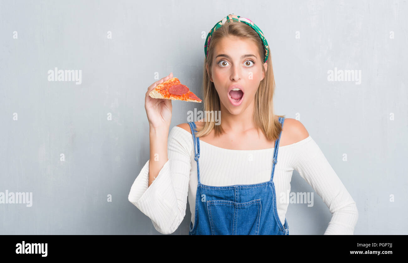 Beautiful young woman over grunge grey wall eating pepperoni pizza ...