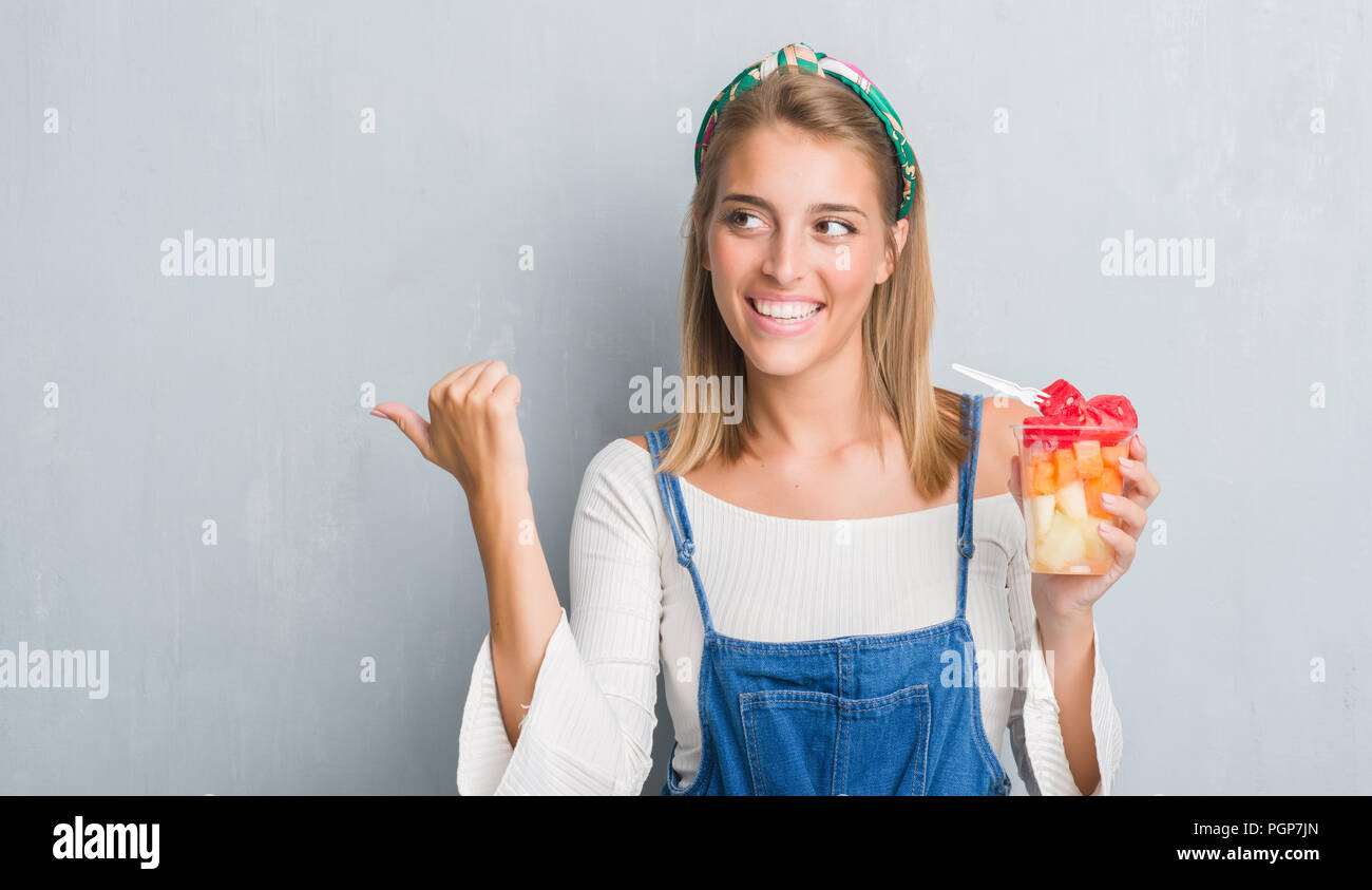 Beautiful young woman over grunge grey wall eating fruits pointing and ...