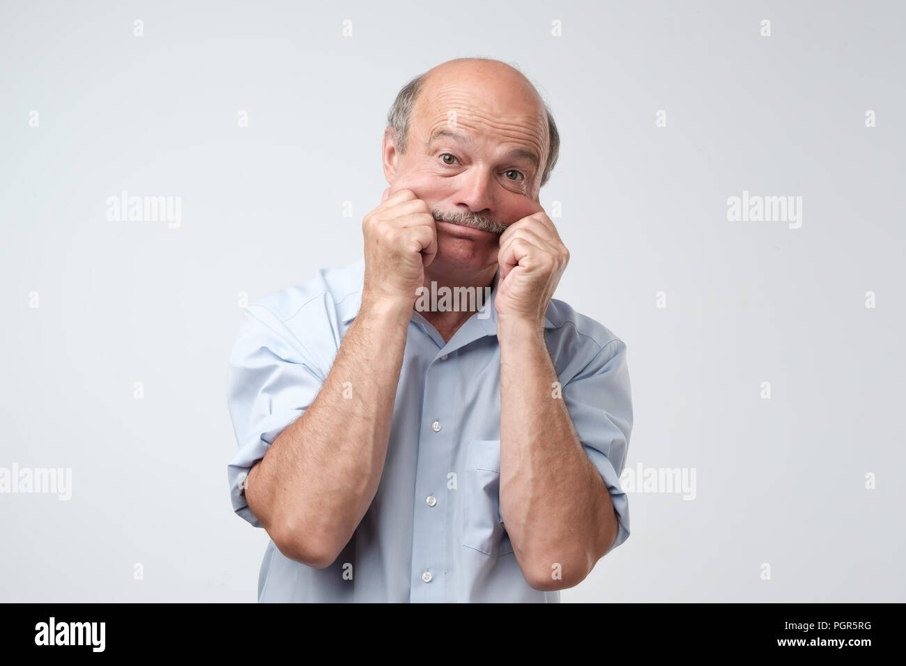 Senior hispanic man in blue shirt pulling skin on face with hands. He ...