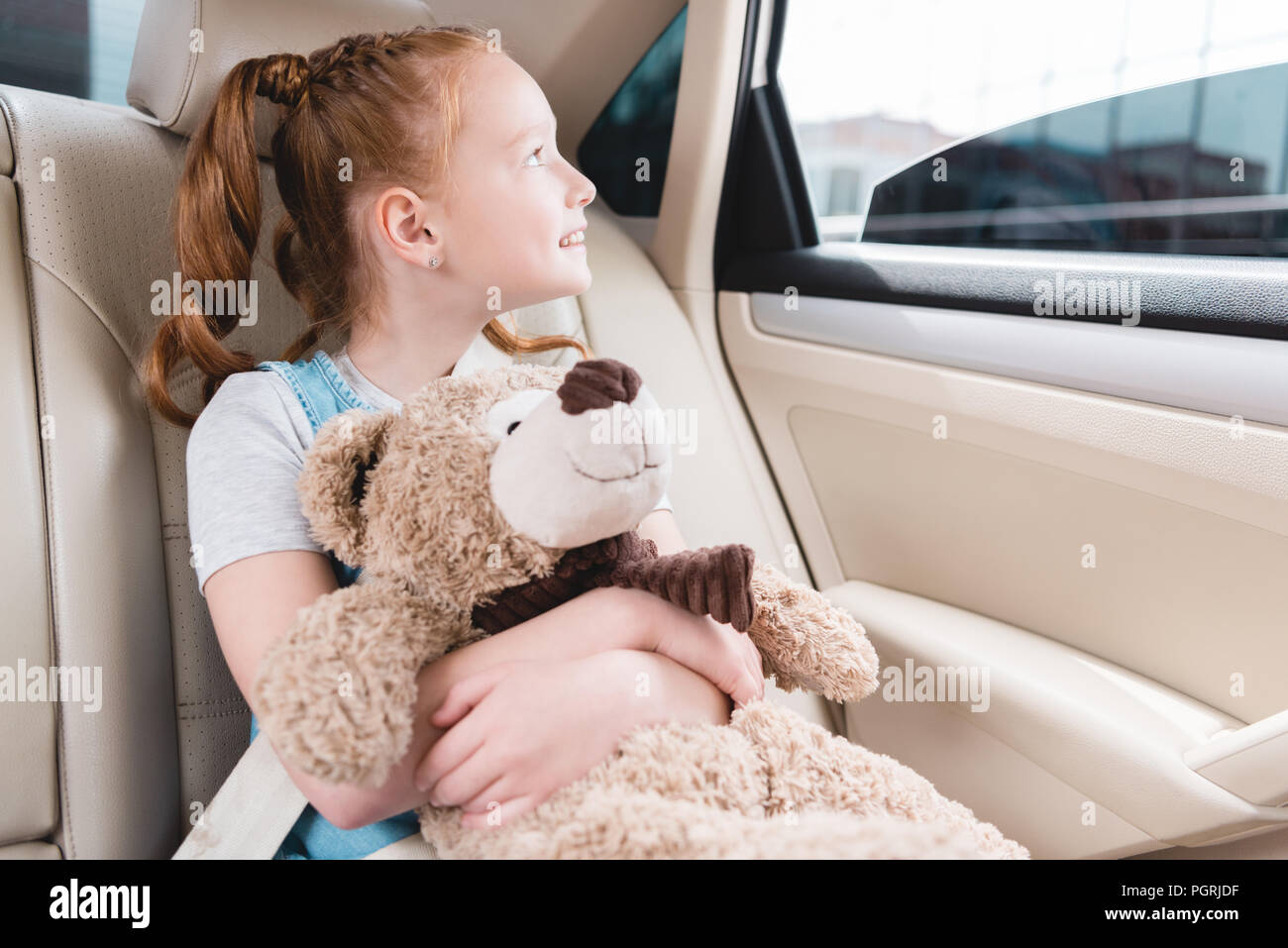portrait of cheerful kid hugging teddy bear and looking out car window ...