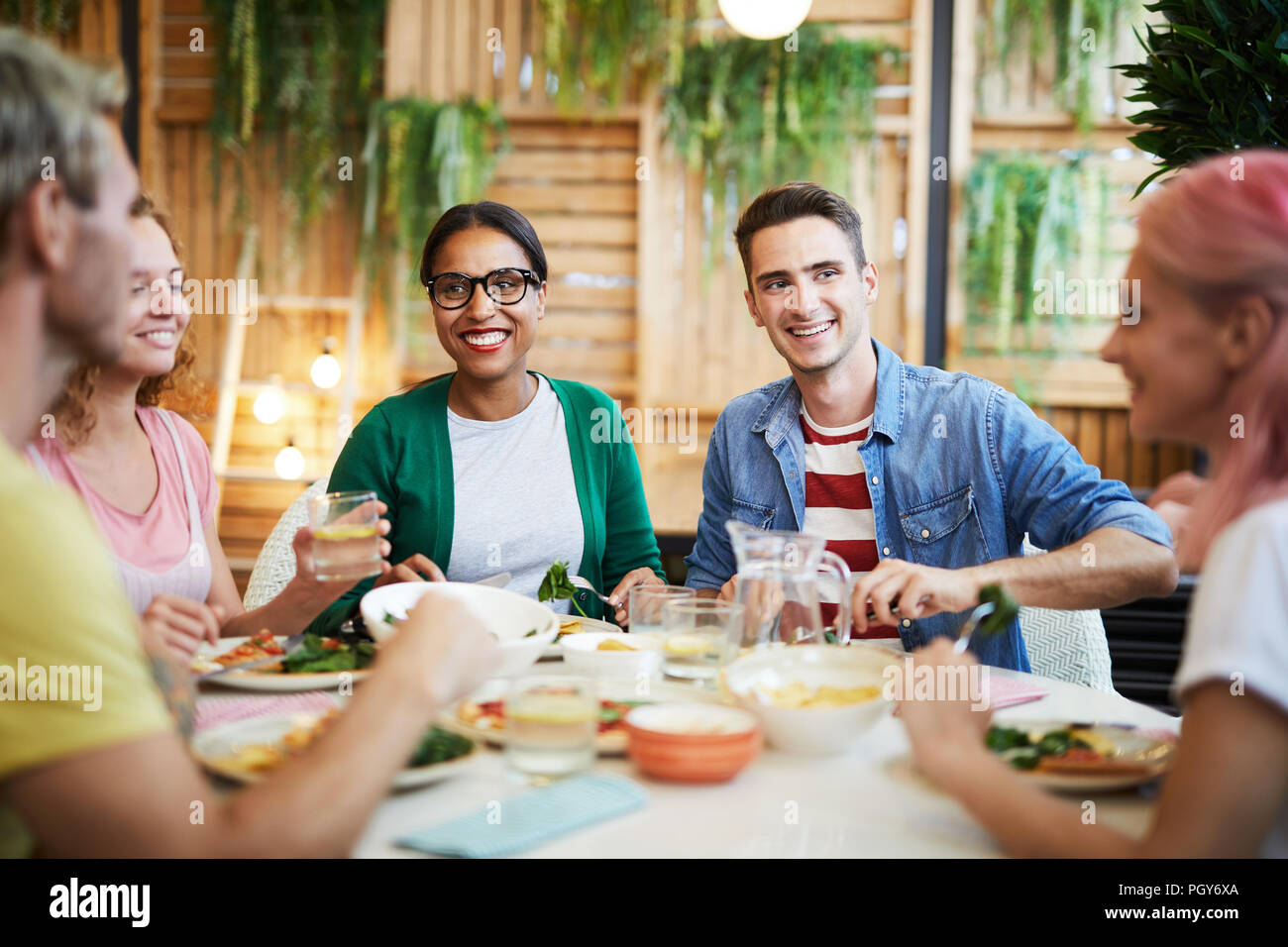 Group of cheerful friends sitting by served table, eating dinner and ...