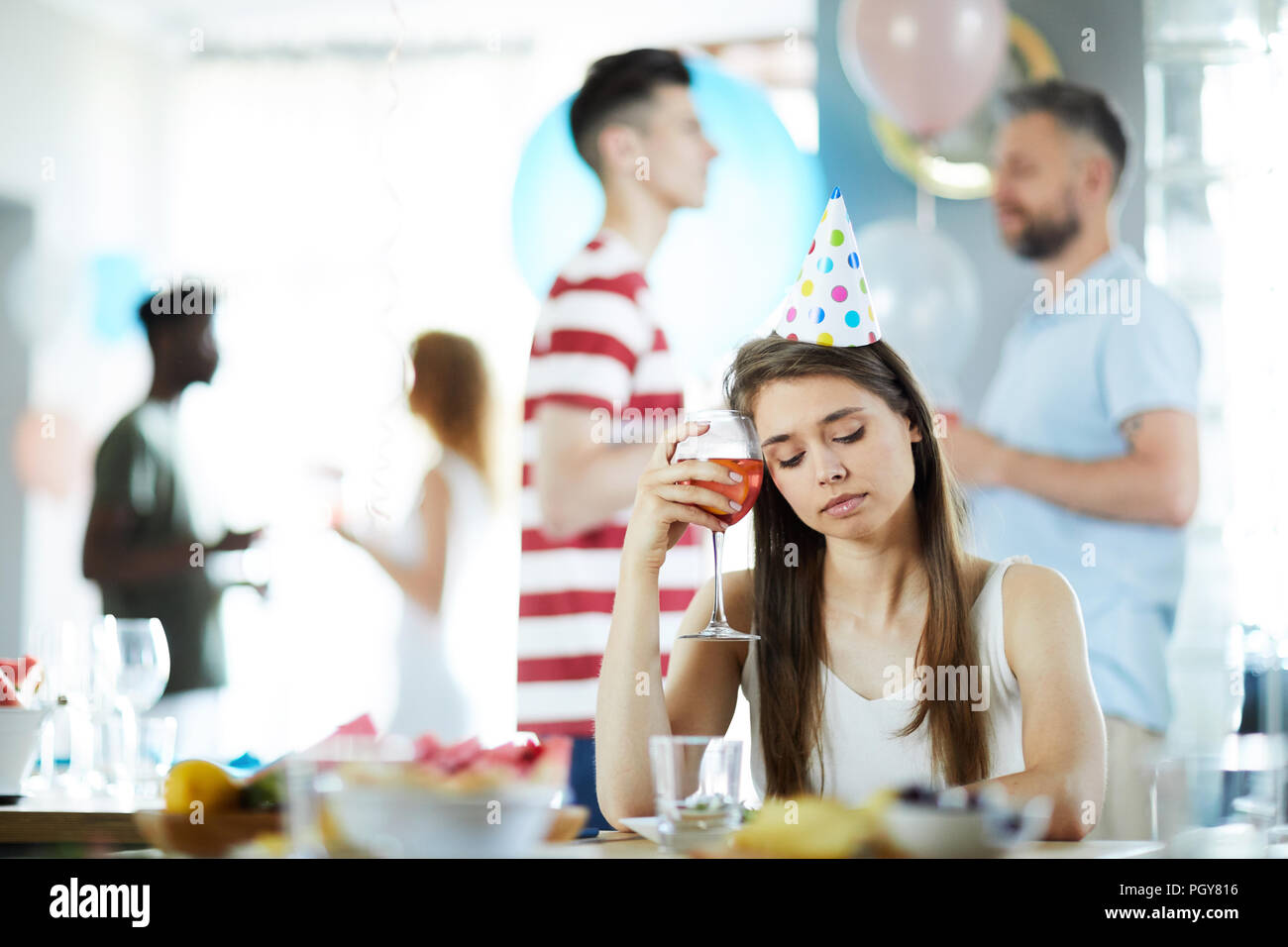 Sad girl in birthday cap sitting by table with drink by her temple on ...