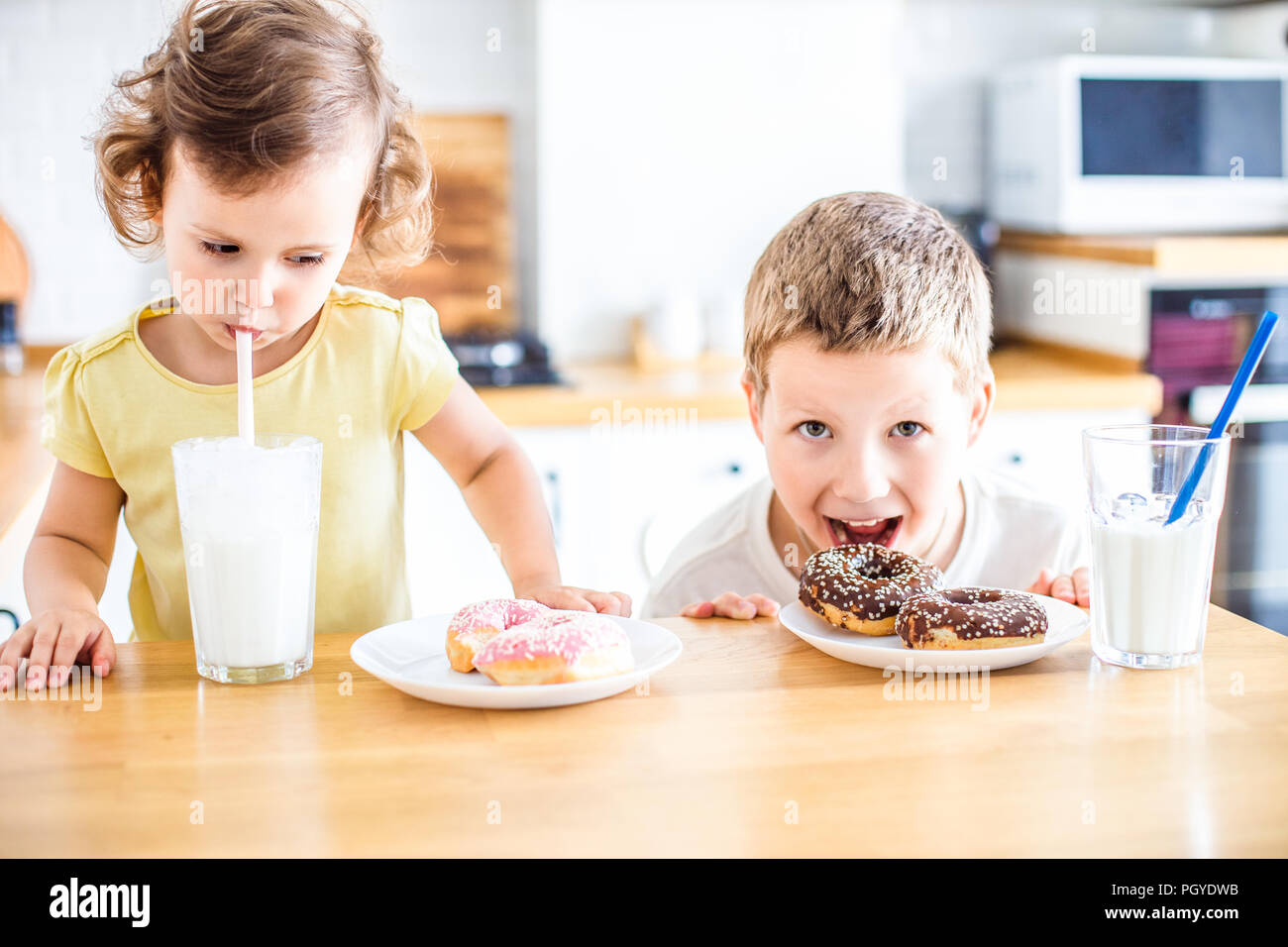 Children eating donuts and drinking milk on the white kitchen at home ...