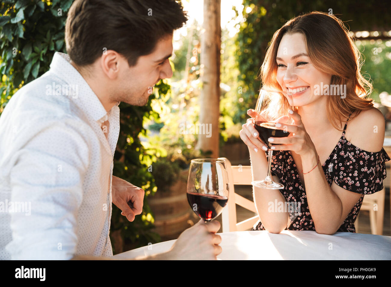 Picture of young loving couple sitting in cafe by dating outdors in park holding glasses of wine ...