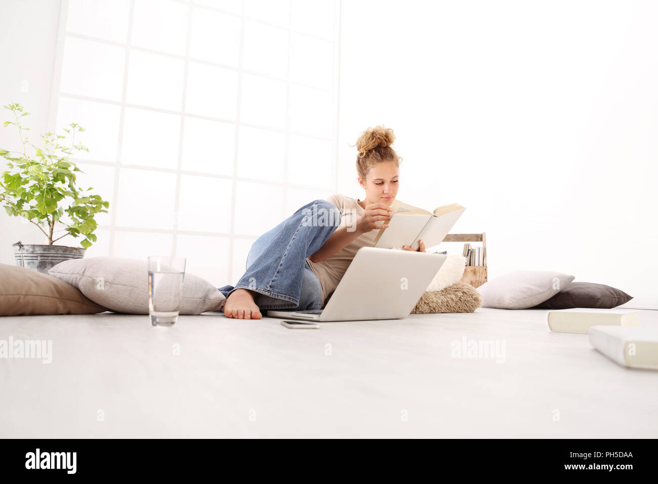 young woman with computer reading a book, lying on the floor in living ...