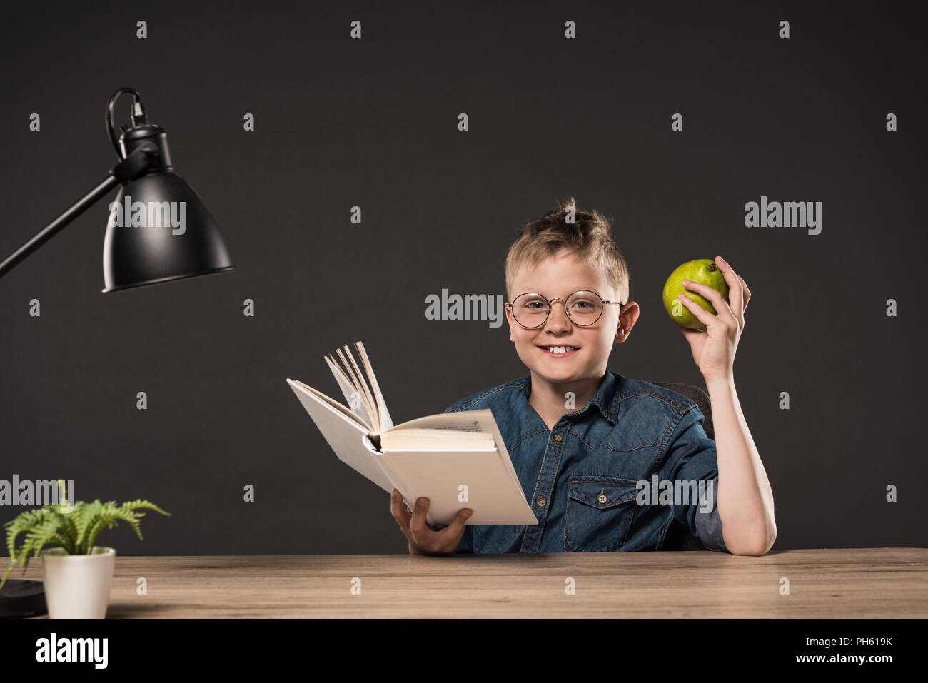 smiling schoolboy holding pear and reading book at table with plant and ...