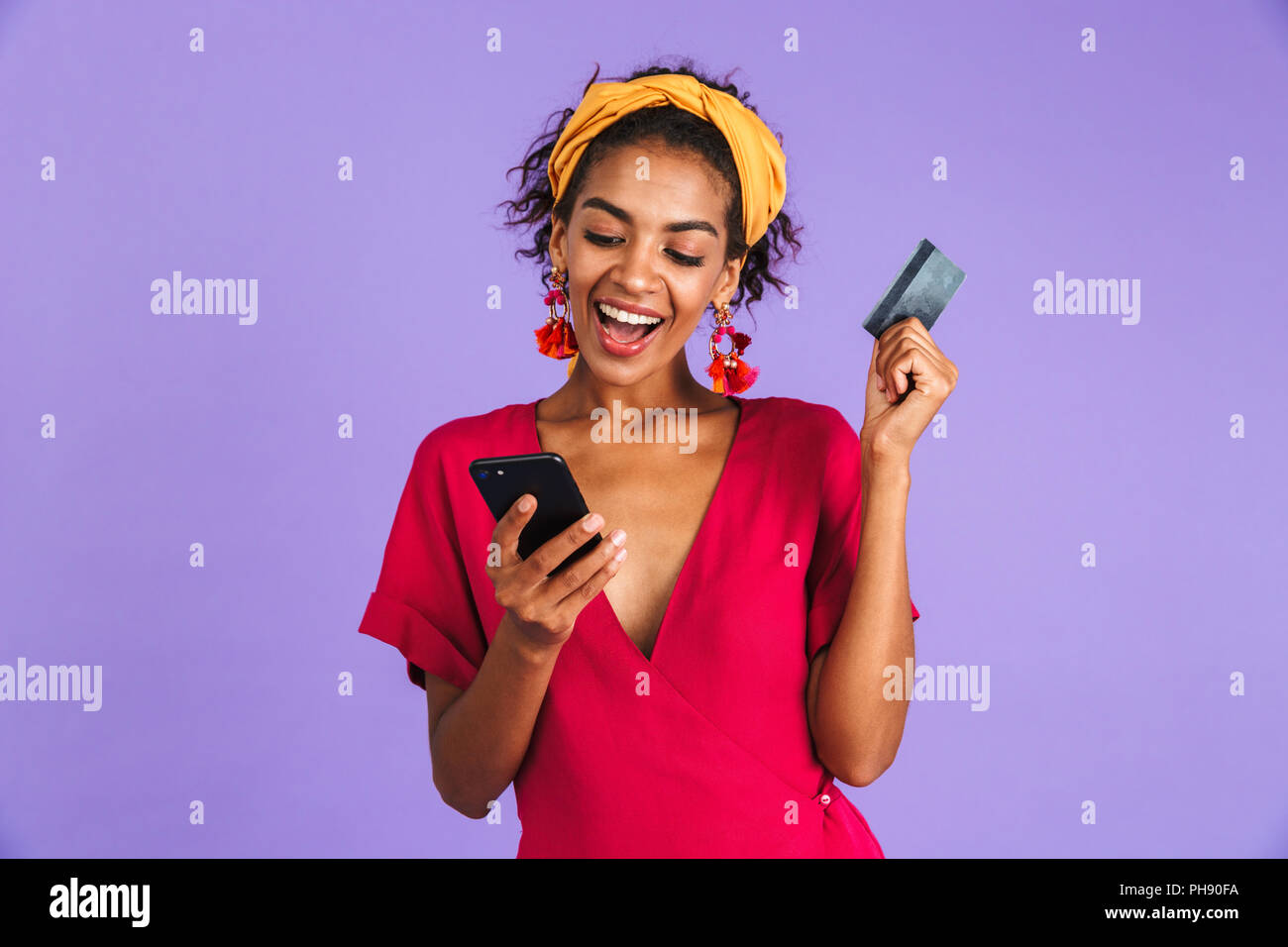 Happy african woman in dress using smartphone while holding credit card over purple background ...
