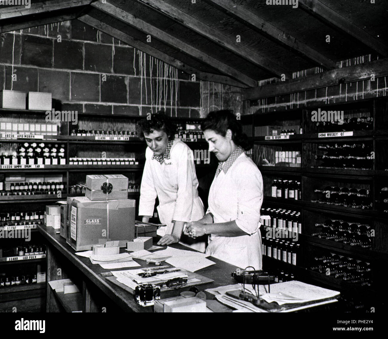 Two women wearing white smocks stand behind the counter of a pharmacy ...
