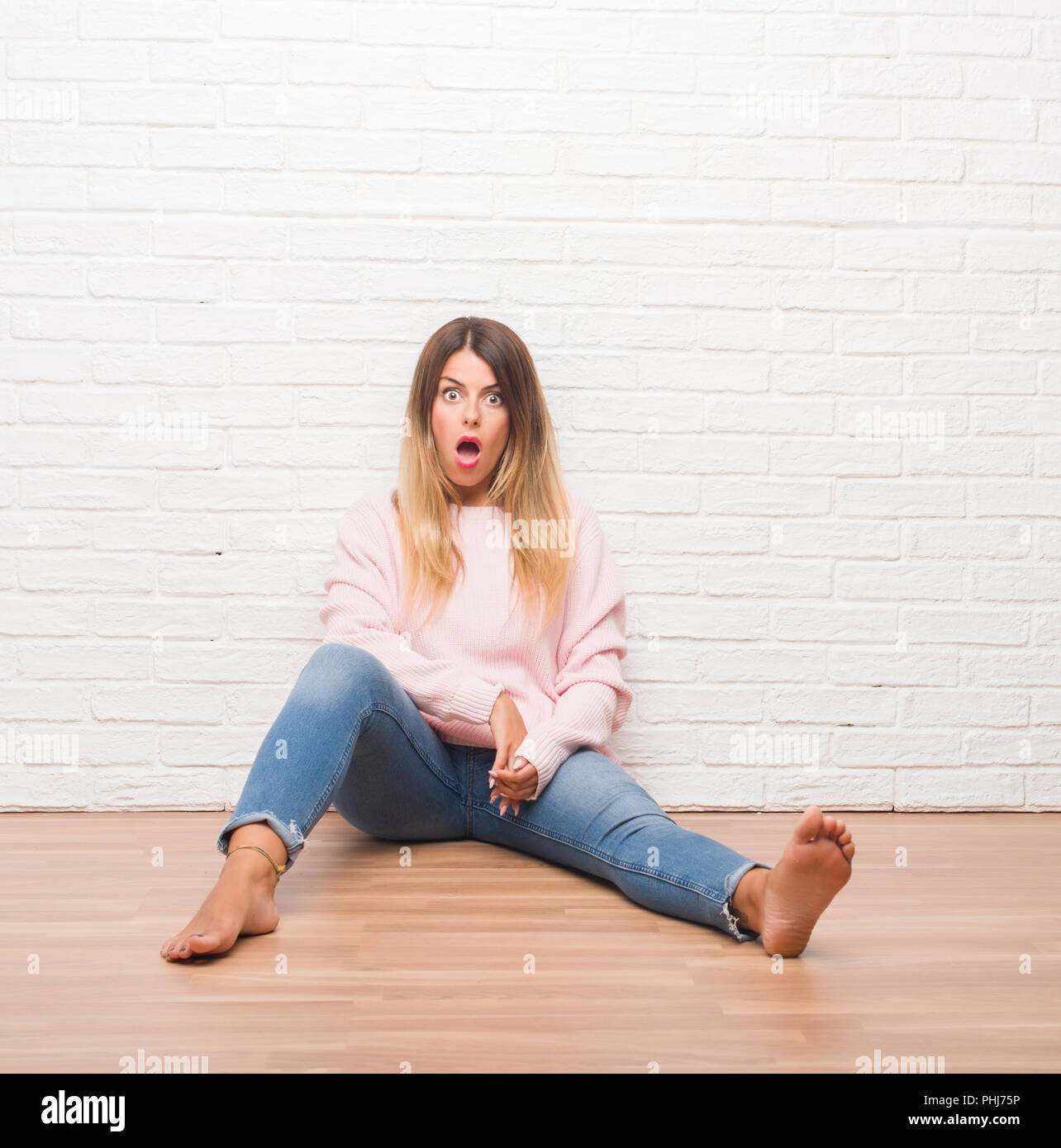 Young adult woman sitting on the floor over white brick wall afraid and ...