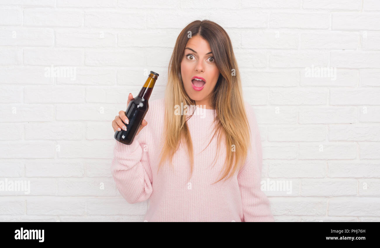Yound adult woman over white brick wall holding beer scared in shock ...