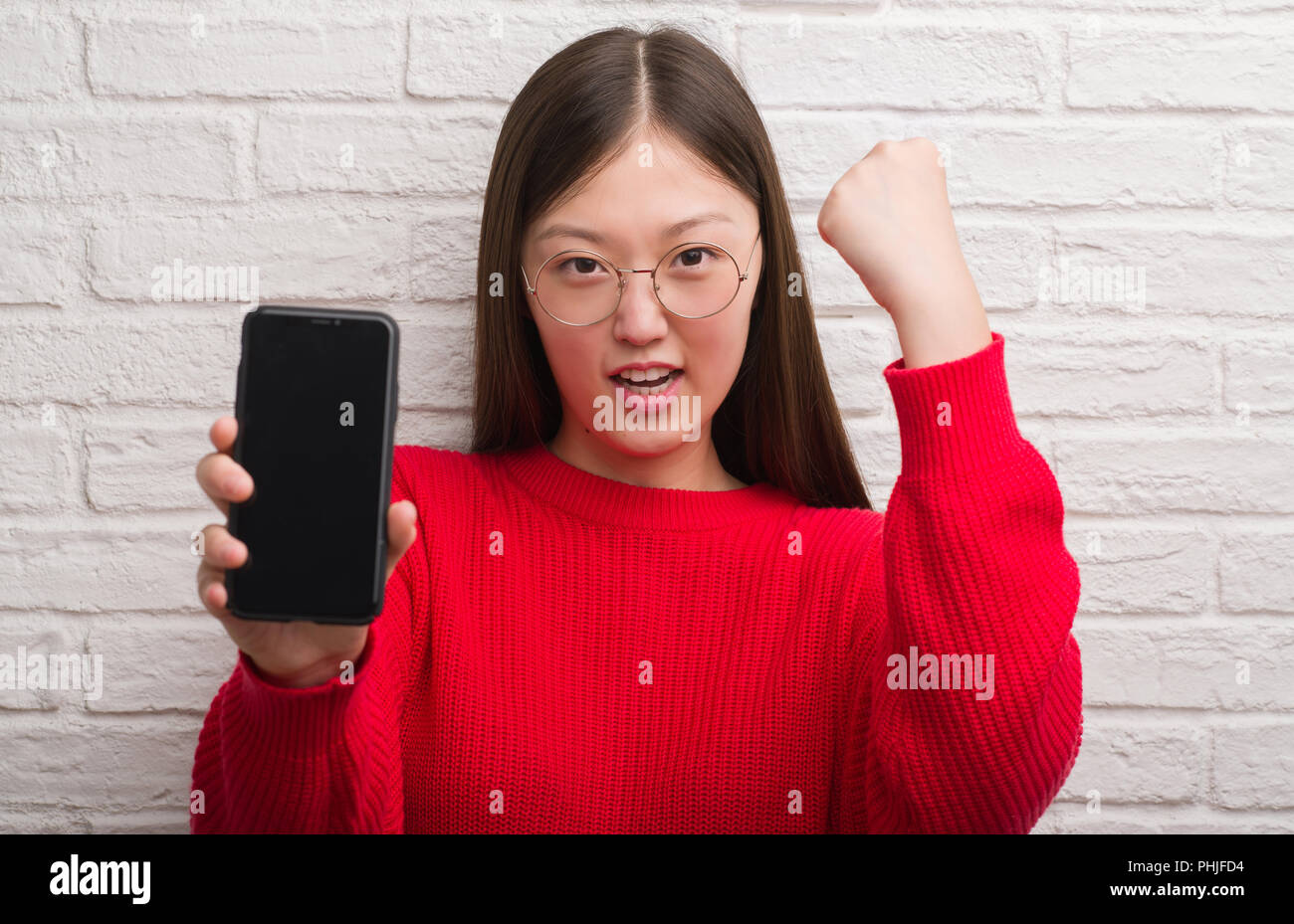 Young Chinese woman over brick wall showing smartphone annoyed and ...