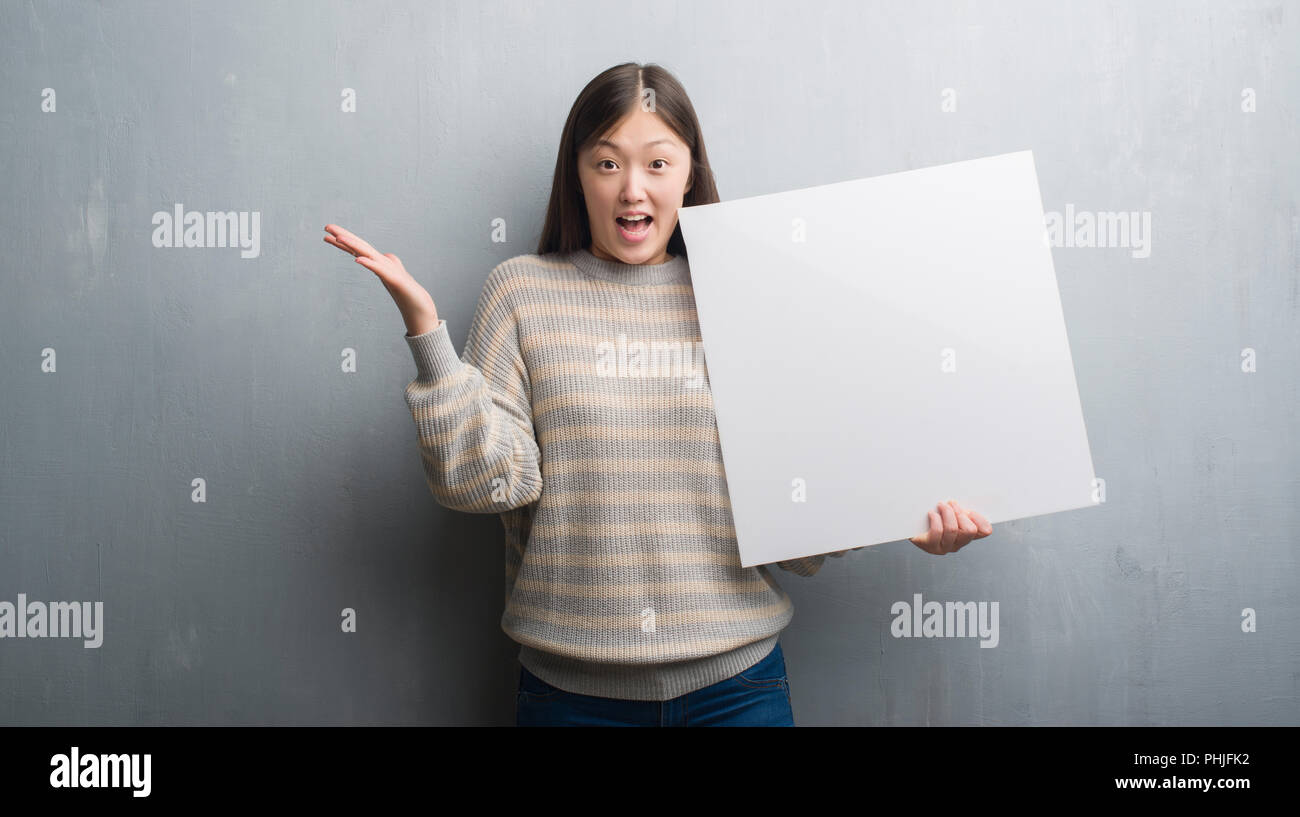 Young Chinese woman over grey wall holding banner very happy and ...