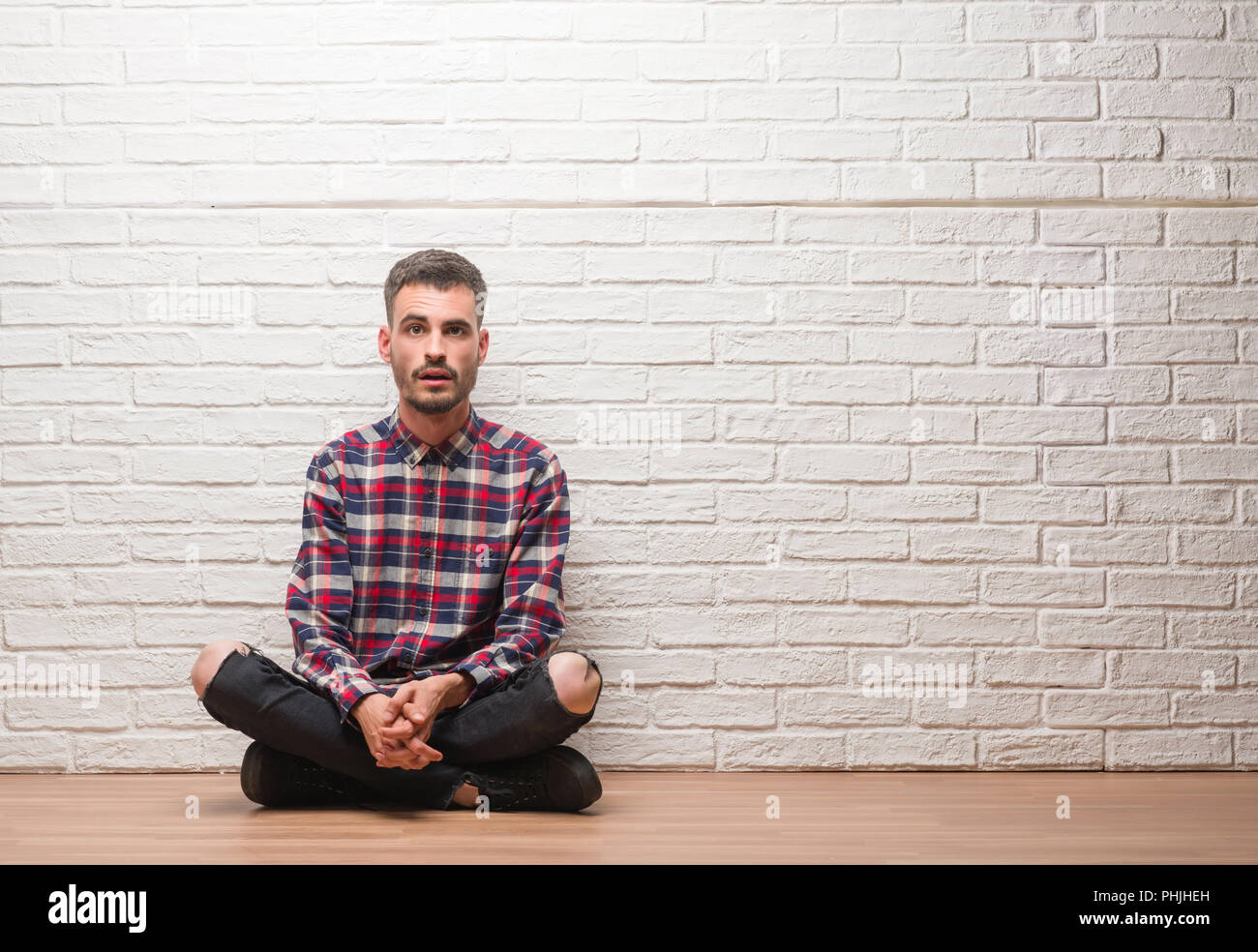 Young adult man sitting over white brick wall scared in shock with a ...