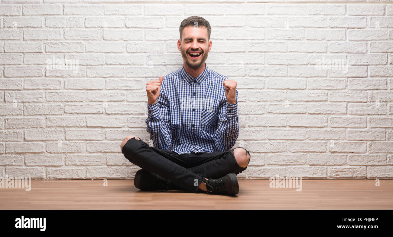 Young adult man sitting over white brick wall excited for success with ...