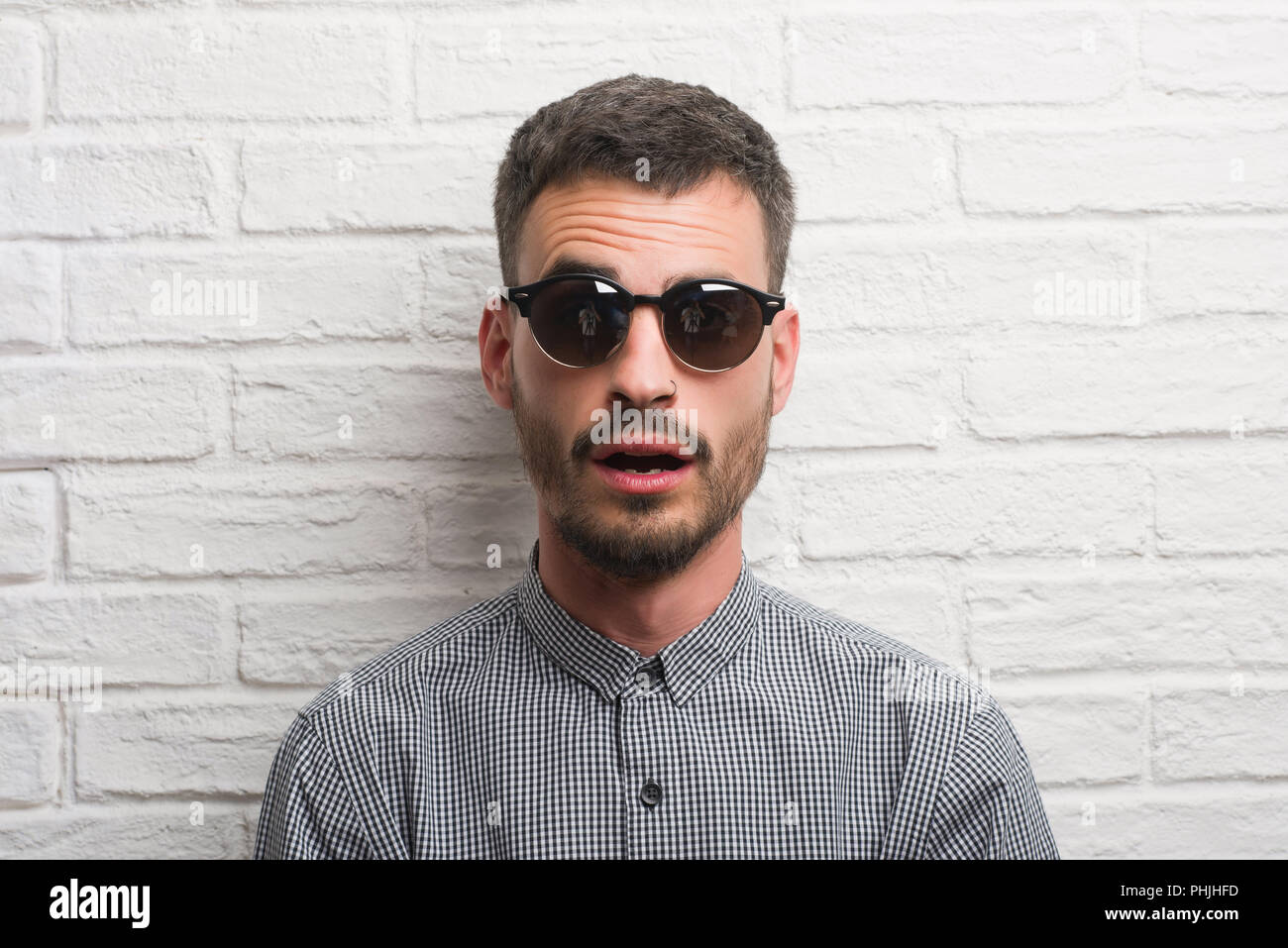 Young adult man wearing sunglasses standing over white brick wall ...