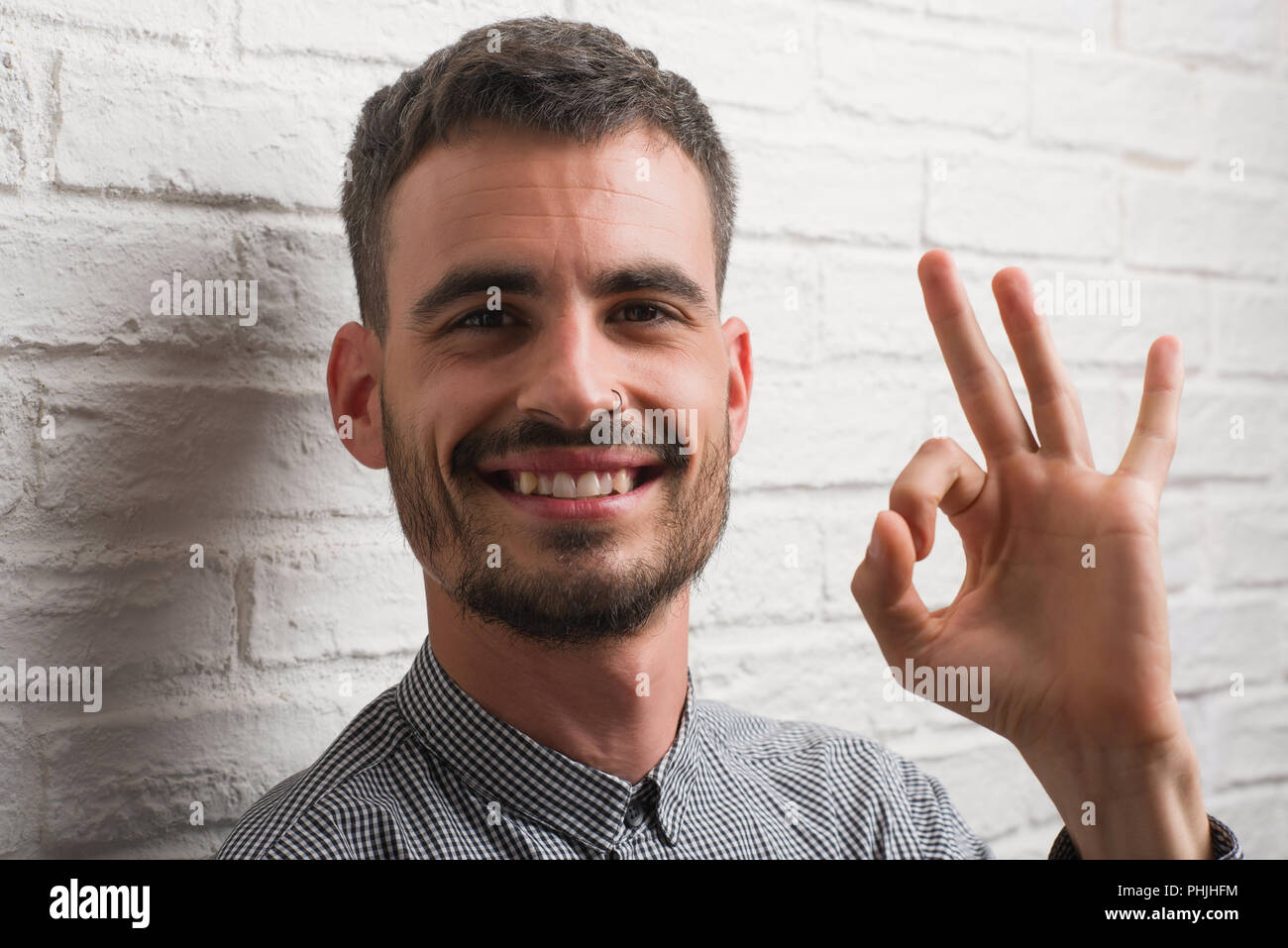 Young adult man standing over white brick wall doing ok sign with ...
