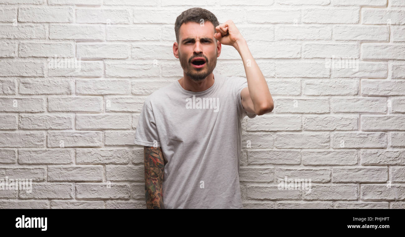 Young adult man standing over white brick wall annoyed and frustrated ...