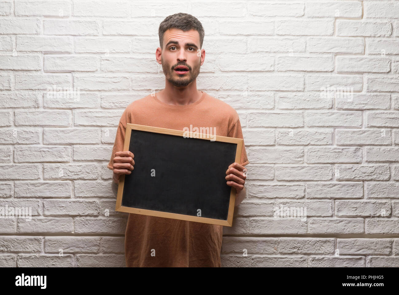 Young adult man over brick wall holding blackboard scared in shock with ...