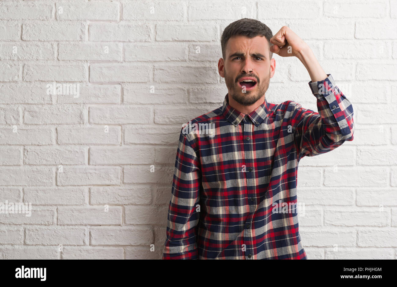 Young adult man standing over white brick wall angry and mad raising ...