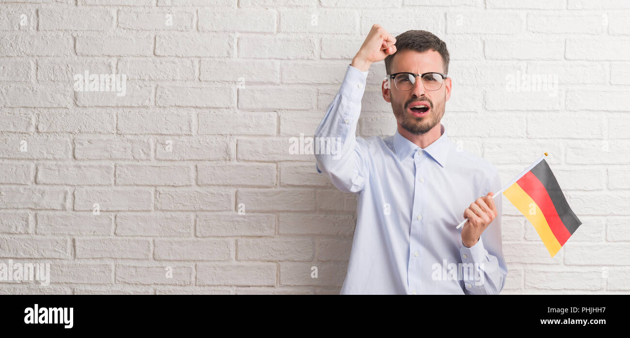 Young adult man over brick wall holding flag of Germany annoyed and ...