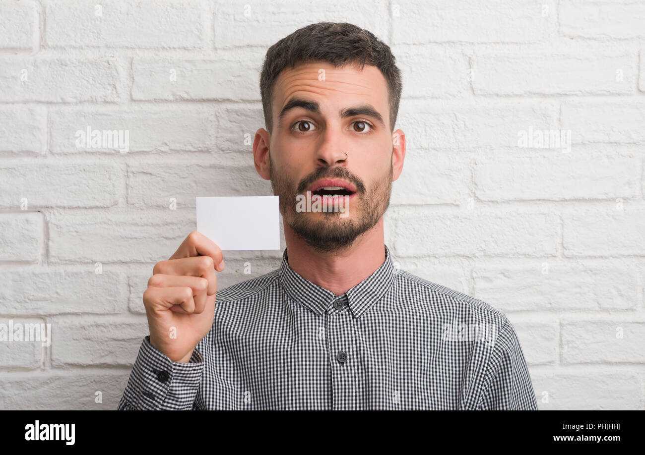 Young adult man over brick wall holding blank card scared in shock with ...
