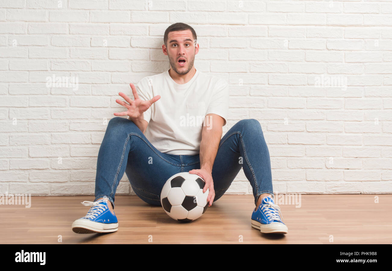 Young caucasian man sitting over white brick wall holding soccer ...