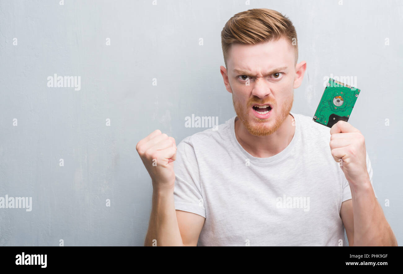 Young redhead man over grey grunge wall holding hard drive annoyed and ...