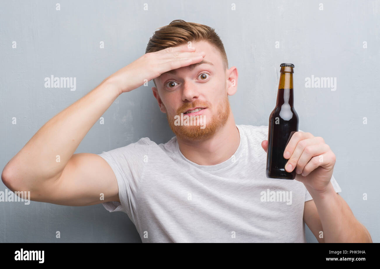 Young redhead man over grey grunge wall drinking beer bottle stressed ...