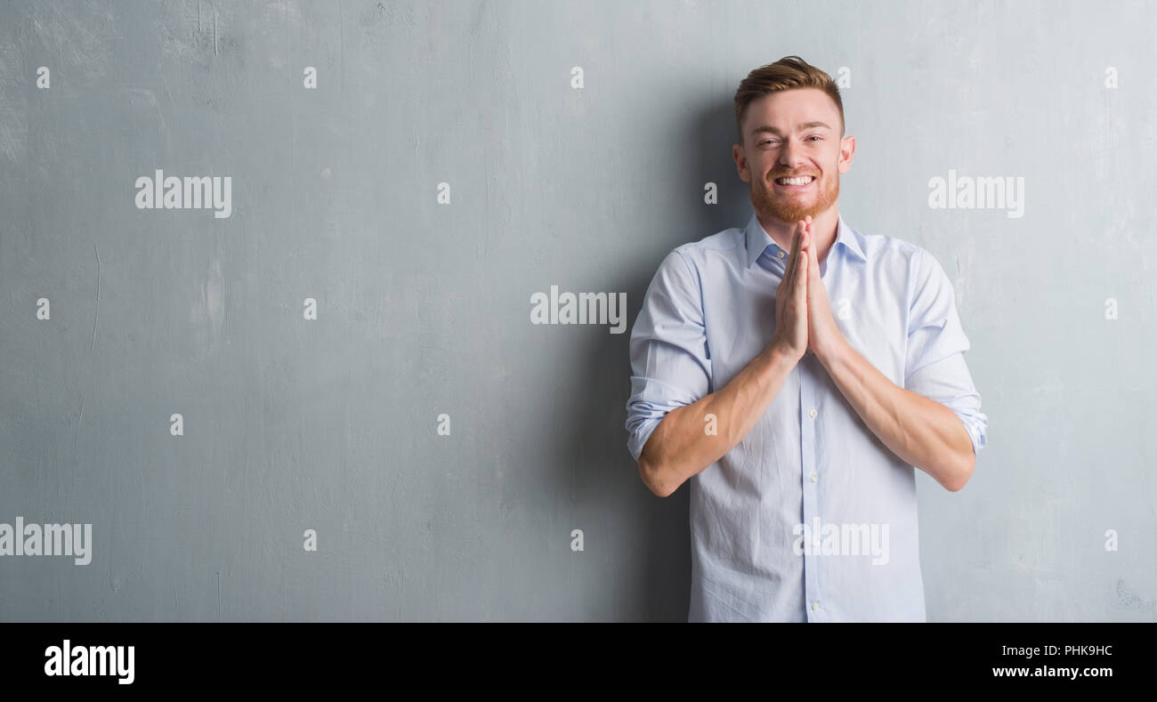 Young redhead business man over grey grunge wall praying with hands ...