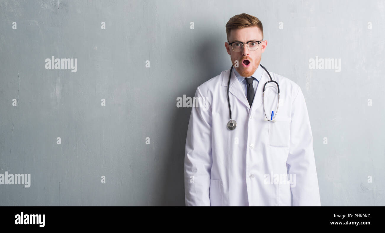 Young redhead doctor man over grey grunge wall scared in shock with a ...
