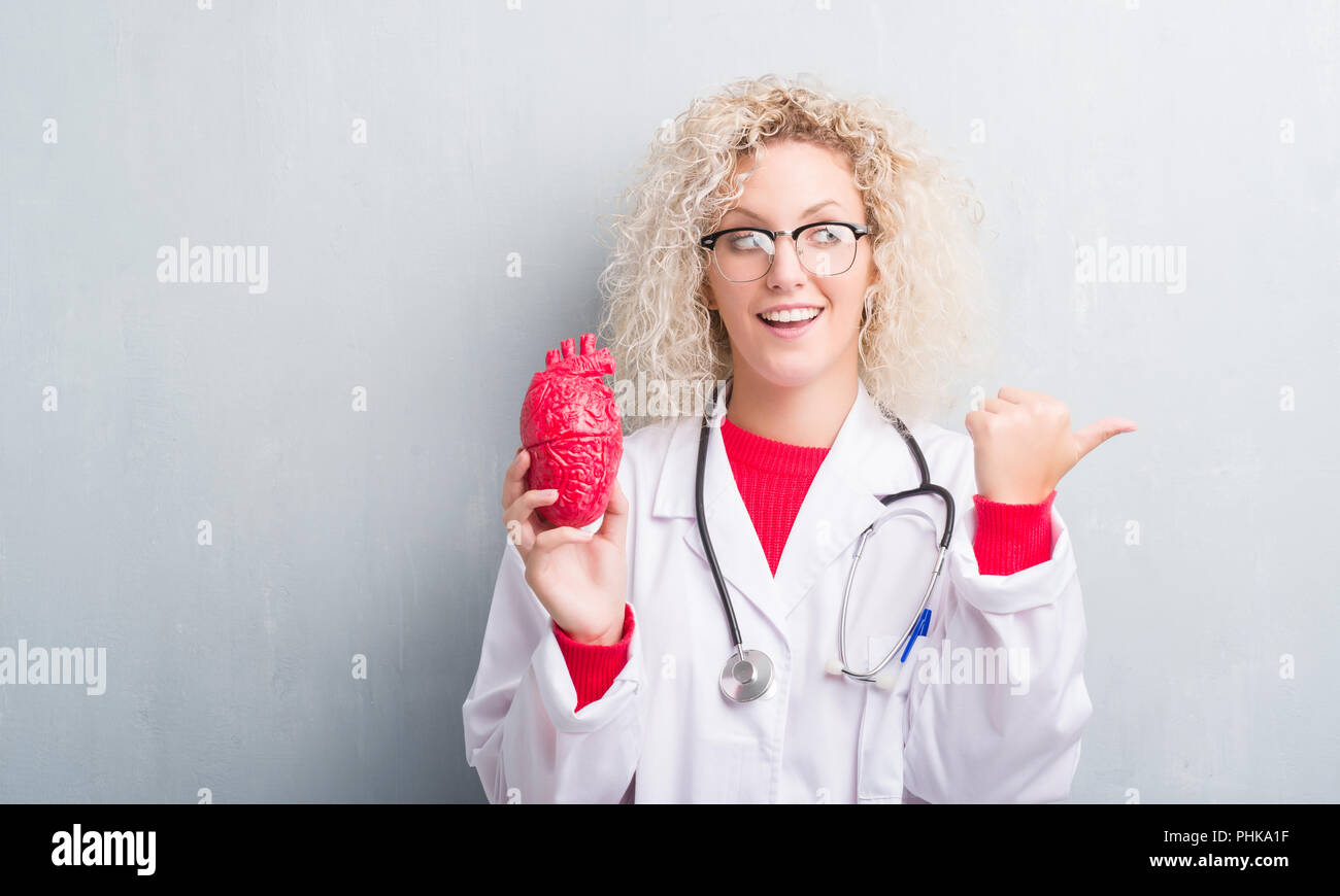 Young blonde cardiologist doctor woman over grunge grey wall holding ...