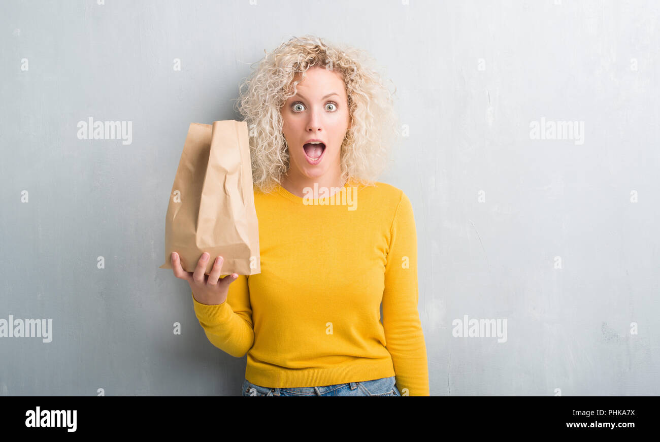 Young blonde woman over grunge grey background holding lunch paper bag ...