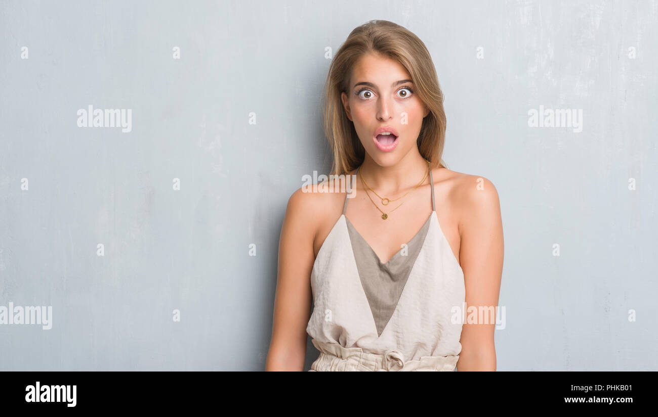 Beautiful young woman standing over grunge grey wall scared in shock ...