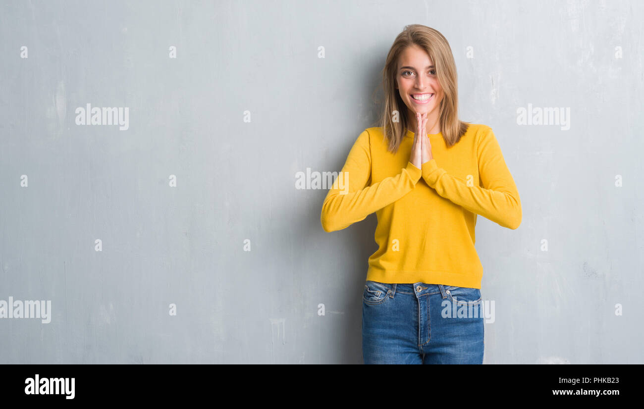 Beautiful young woman standing over grunge grey wall praying with hands ...