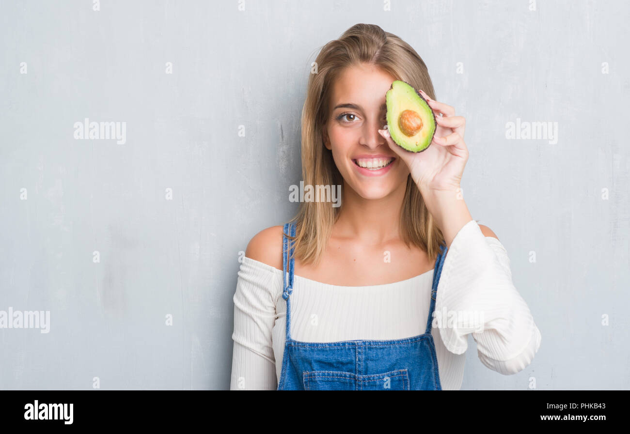 Beautiful young woman over grunge grey wall eating avocado with a happy ...