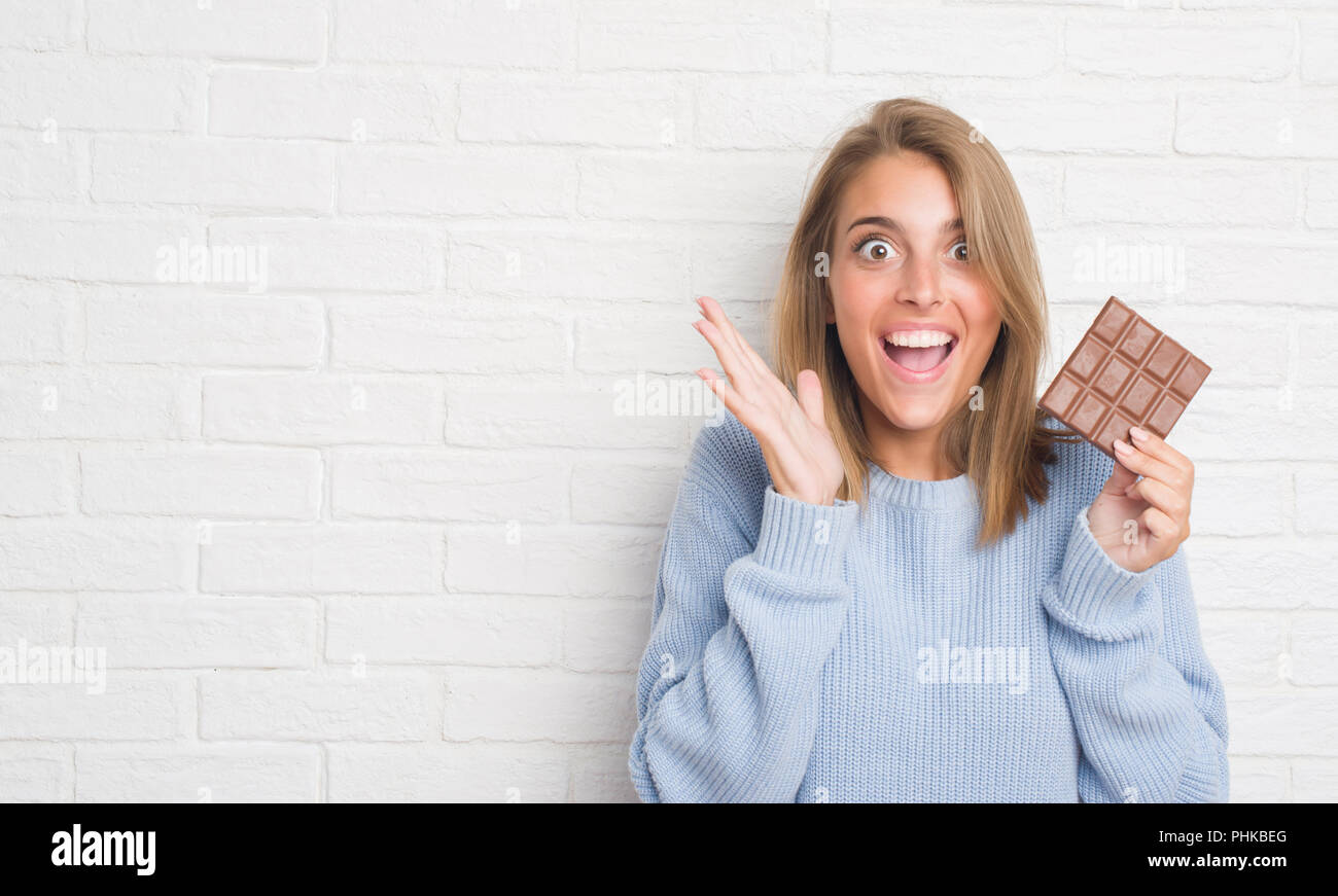 Beautiful young woman over white brick wall eating chocolate bar very ...