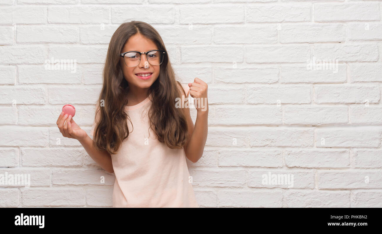 Young hispanic kid over white brick wall eating pink macaron screaming ...