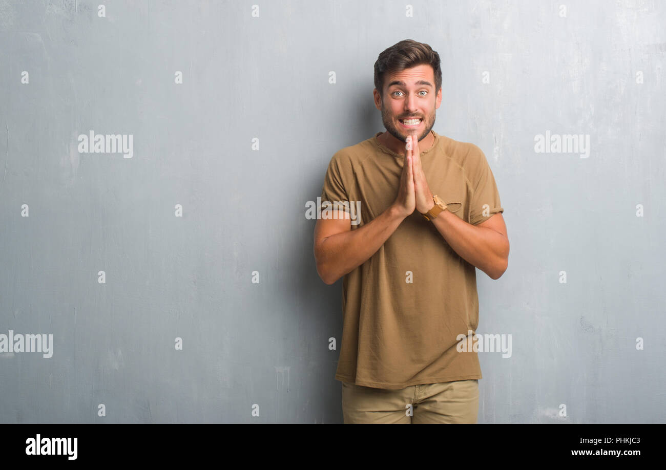 Handsome young man over grey grunge wall praying with hands together ...