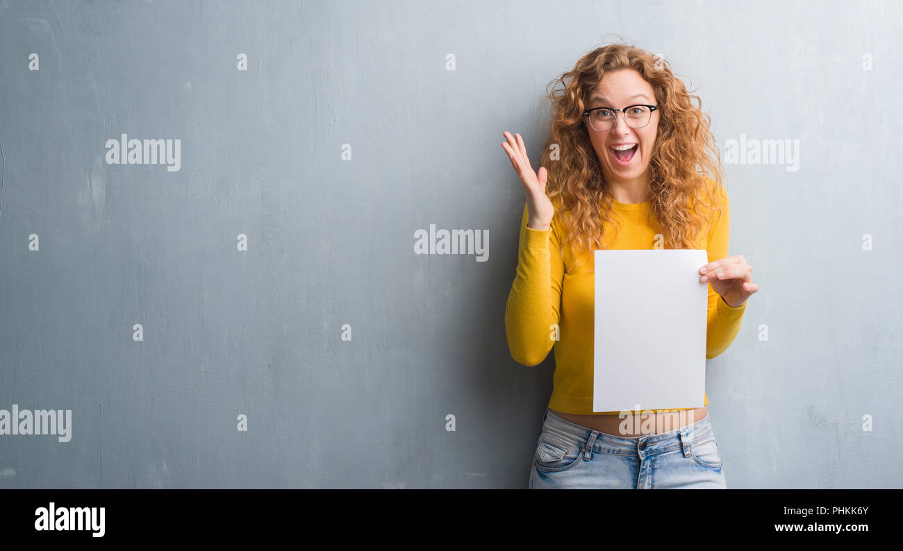 Young redhead woman over grey grunge wall holding blank paper sheet ...