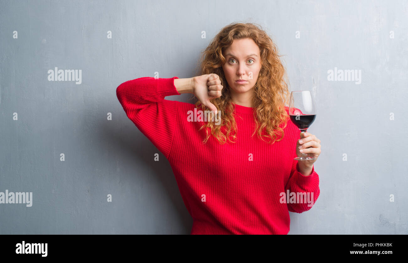 Young redhead woman over grey grunge wall drinking a glass of wine with ...