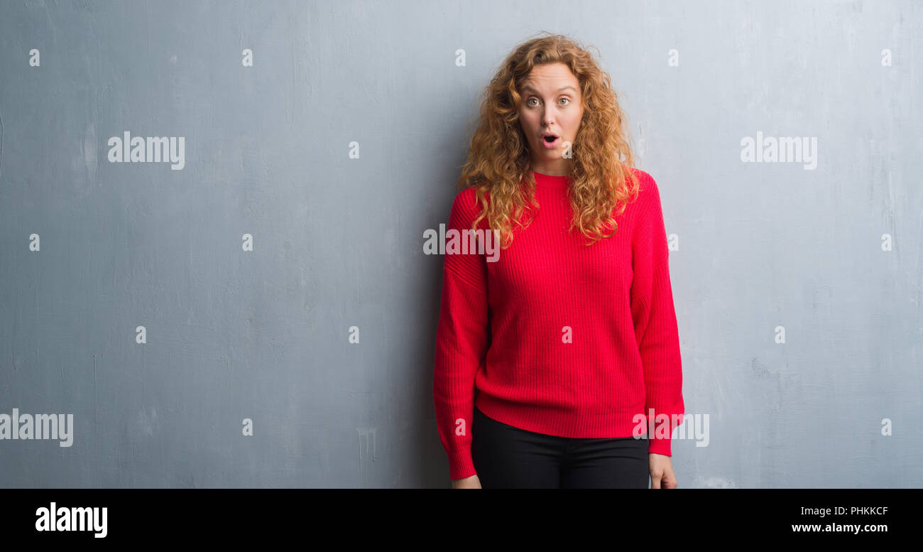Young redhead woman over grey grunge wall wearing red sweater afraid ...